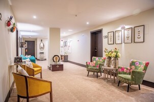Um saguão de hotel com carpete bege, poltronas florais em amarelo e verde, um sofá azul e uma exibição emoldurada de borboletas na parede.