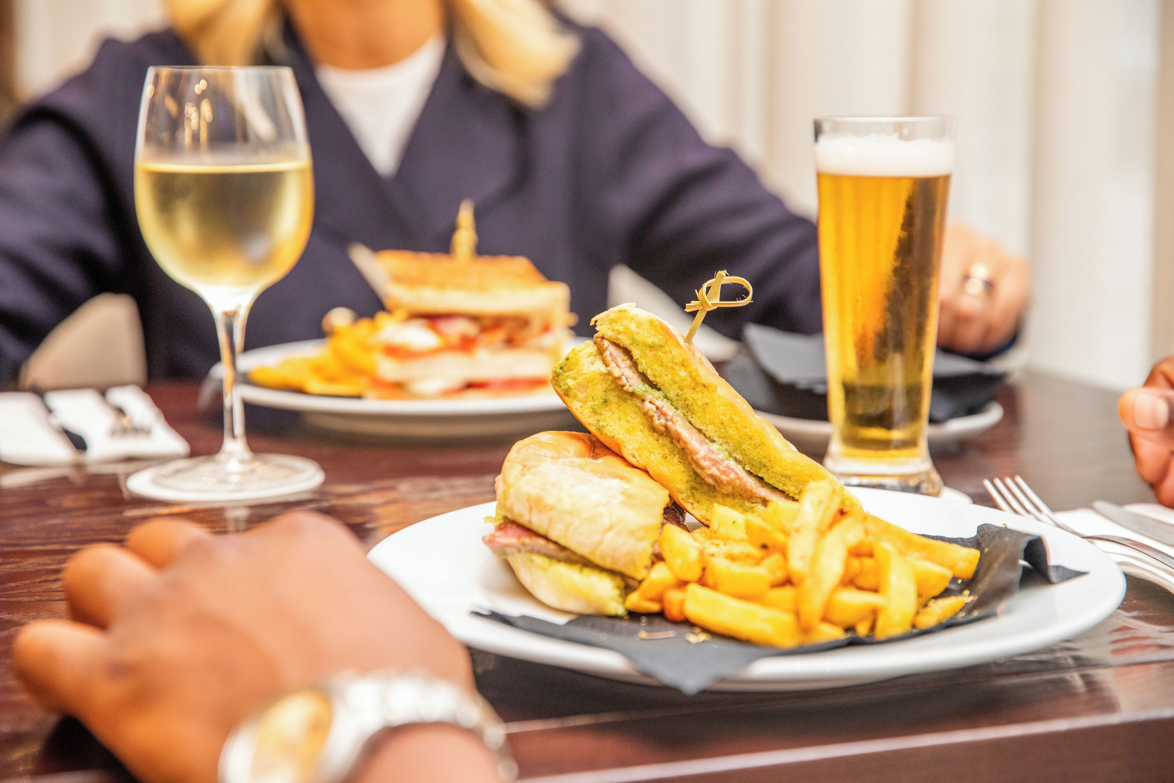 Uma refeição com sanduíche e batatas fritas em um prato, uma caneca de cerveja e um copo de vinho branco.