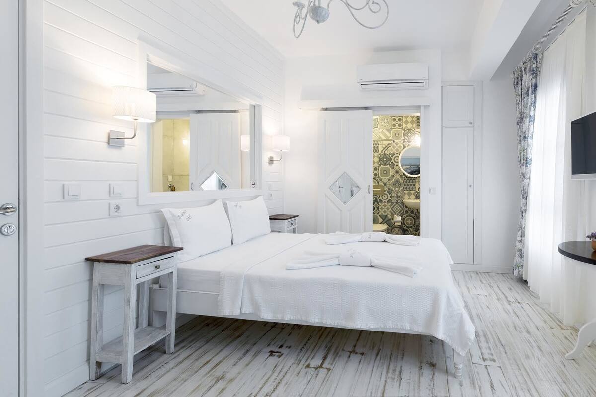 Quarto com decoração em tons de branco, incluindo cama, mesas de cabeceira, espelho, ar condicionado, televisão e vista para o exterior pela janela.