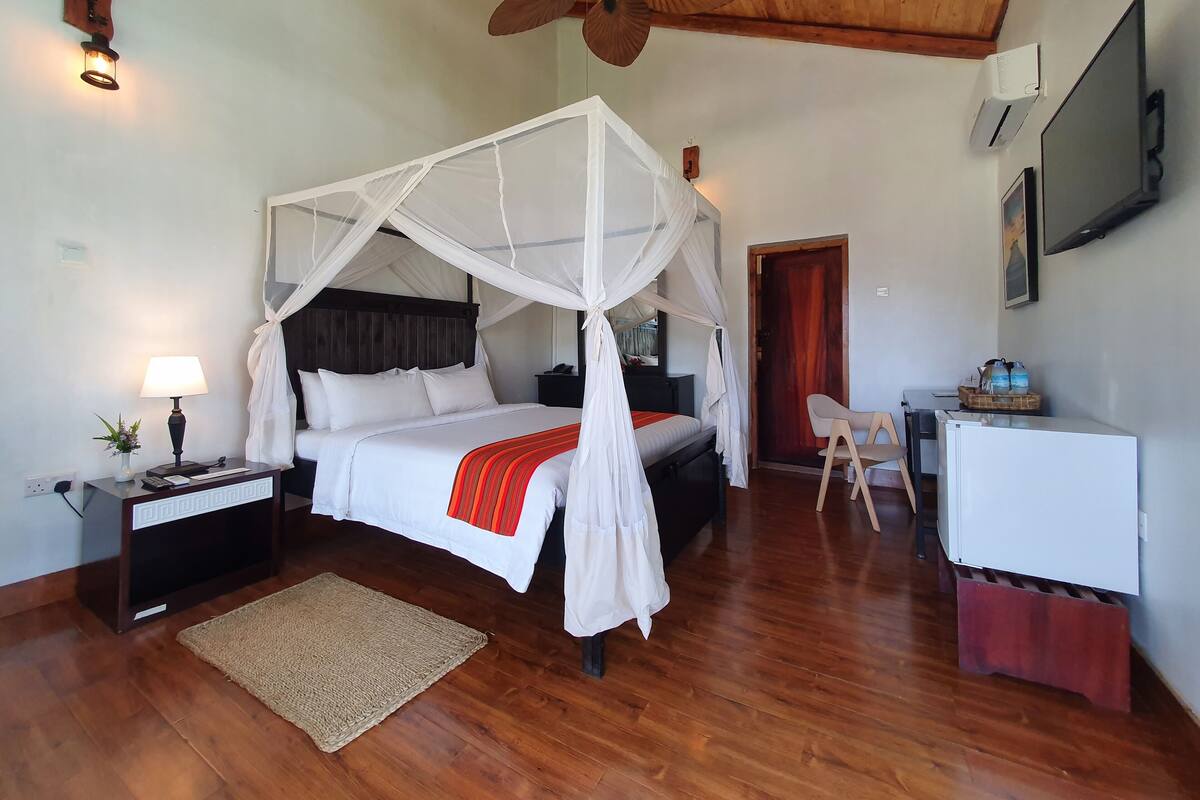 Quarto com cama de dossel, escrivaninha com cadeira, ventilador de teto e televisão.