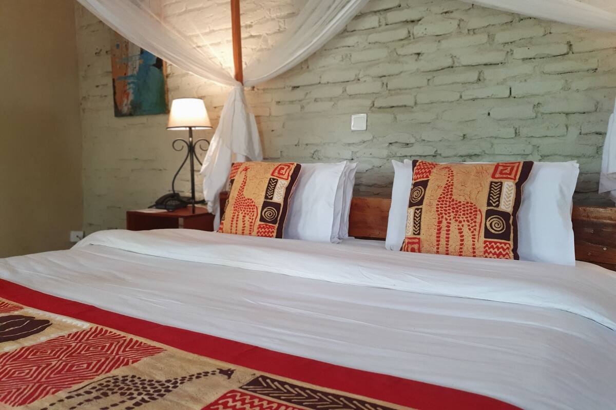 Uma cama com lençóis brancos e almofadas estampadas, uma mesa de cabeceira com abajur e uma parede de tijolos.