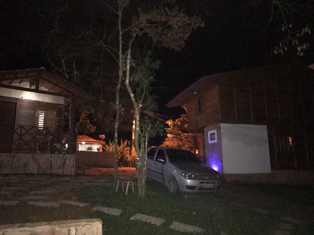 Vista noturna de uma área residencial com um carro estacionado, uma casa com janela iluminada e uma árvore em primeiro plano.