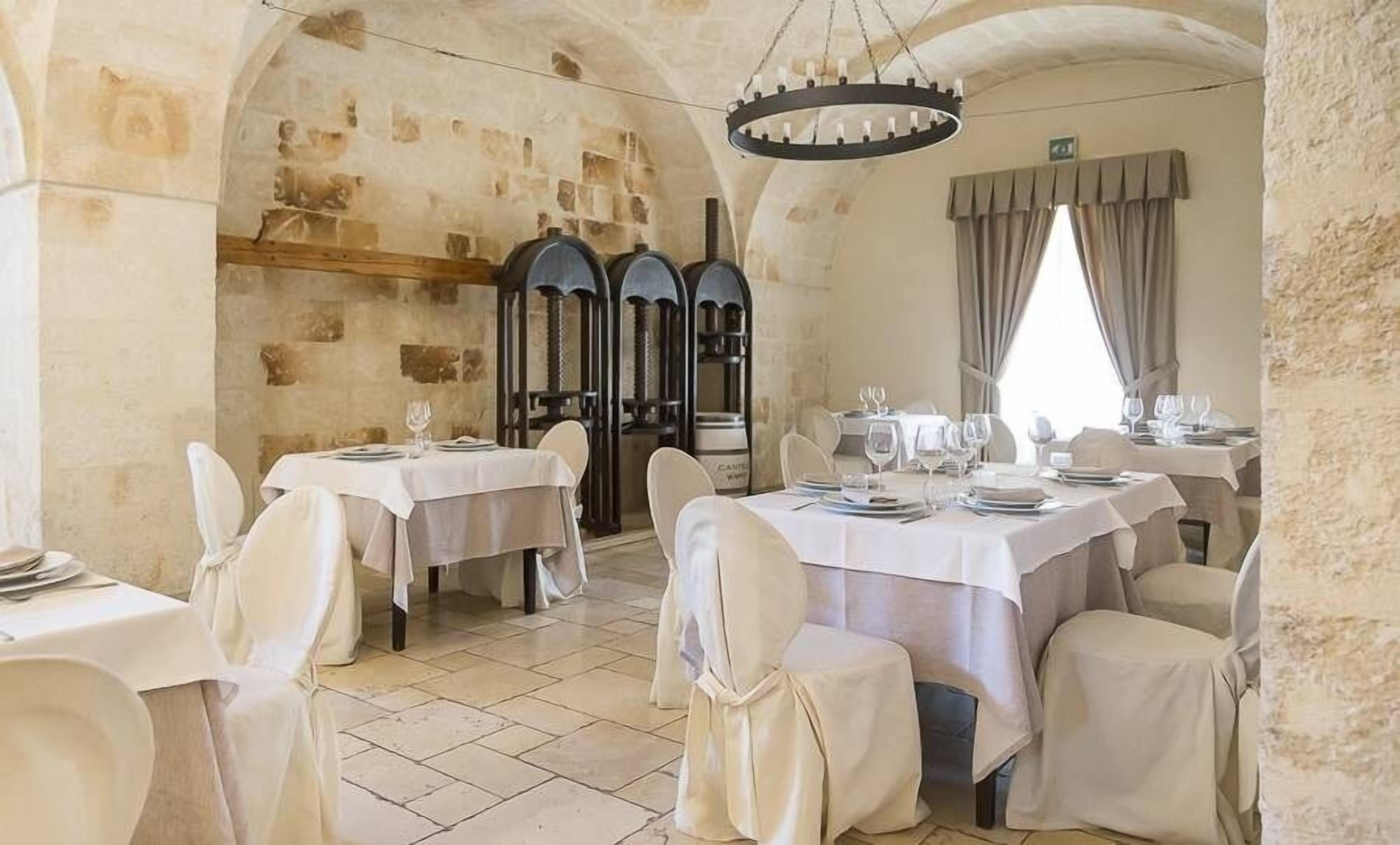Uma área de refeições com paredes de pedra, tetos abobadados e um candelabro. As mesas estão dispostas com toalhas brancas e cadeiras.