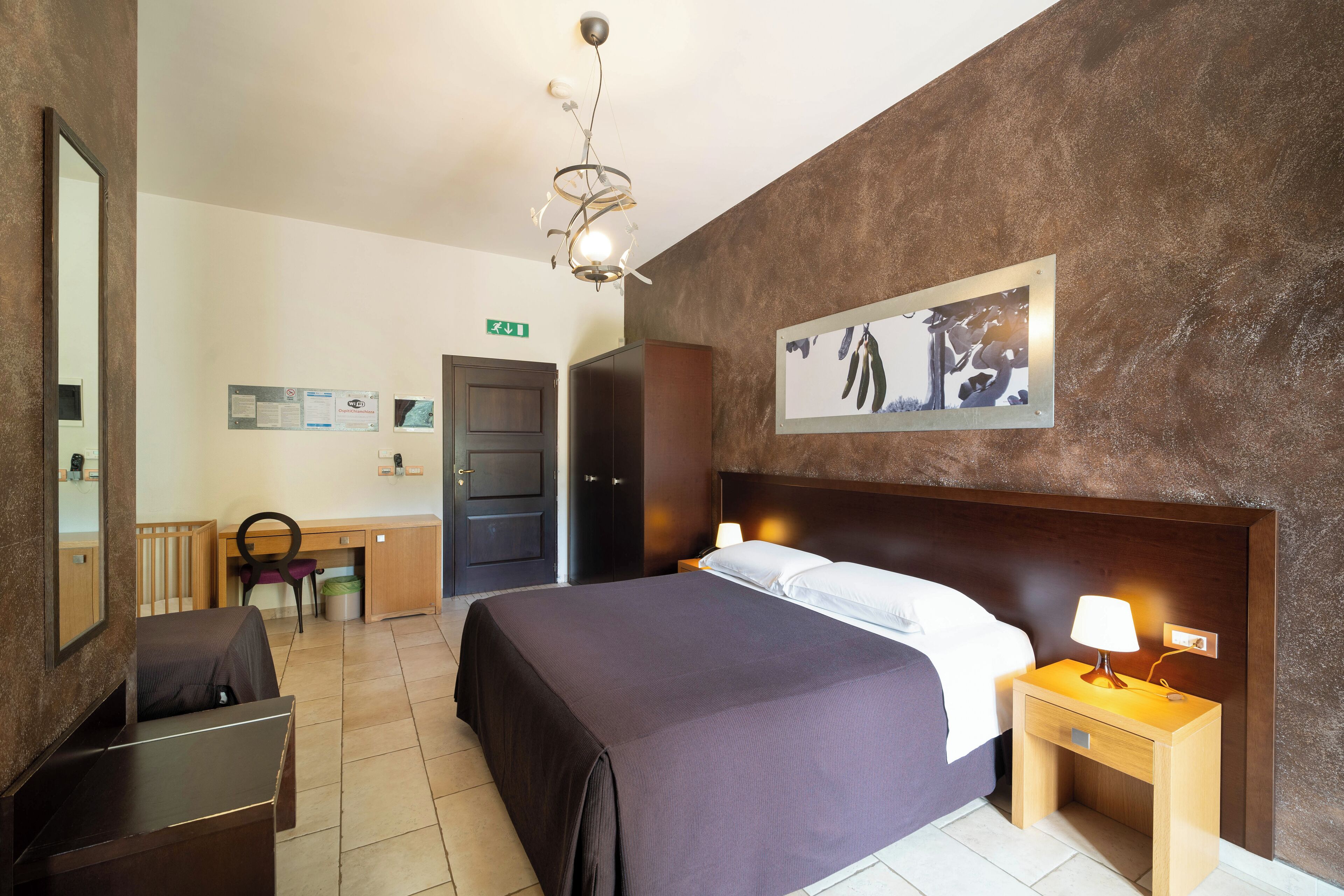 Quarto de hotel com cama, mesa de cabeceira, escrivaninha, cadeira e espelho.