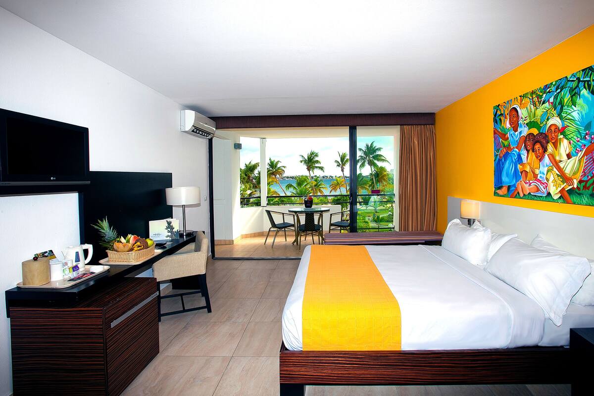 Quarto de hotel com cama, escrivaninha, televisão, varanda e vista para palmeiras e piscina.