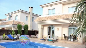 Piscina com espreguiçadeiras e uma bola colorida, cercada por uma casa com telhado telhado e uma varanda.
