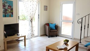 Sala de estar com televisão sobre mesa de madeira, uma cadeira com almofada azul, mesa de centro com um livro e uma planta, e uma porta que dá para o exterior.
