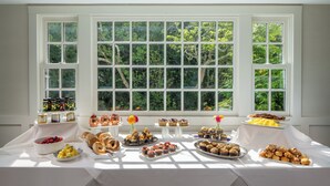 Mesa de buffet de café da manhã com diversos pães e frutas, com vista para um jardim através de grandes janelas.