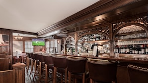 Uma área de bar com banquetas de madeira, uma tela de televisão e prateleiras com garrafas e copos.