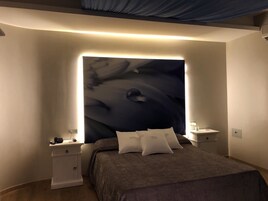 Quarto de hotel moderno com uma cama grande, uma mesinha de cabeceira com telefone e luminária, e uma cabeceira fixada na parede com iluminação ambiente.