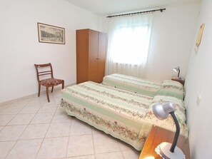 Um quarto com cama, uma cadeira, guarda-roupa, escrivaninha com luminária e janela com cortinas.