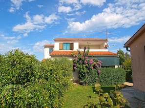 Uma casa de dois andares com telhado de telhas vermelhas, varanda e jardim com arbustos verdes e flores cor-de-rosa.