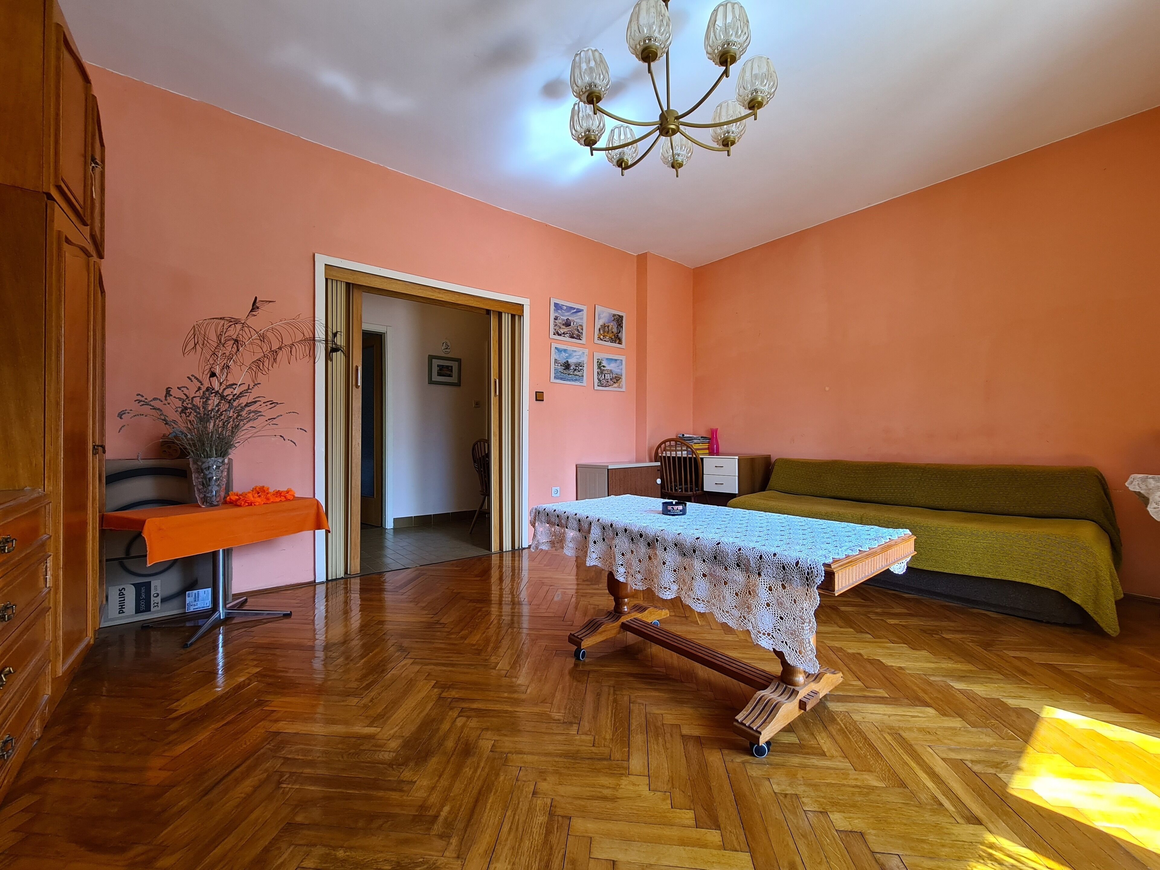 Um quarto com piso de madeira, uma mesa com toalha rendada, um sofá verde, um armário de madeira e um lustre.