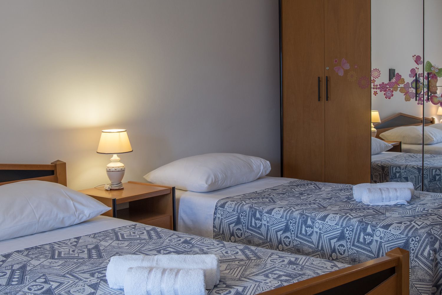Quarto de hotel com duas camas, mesa de cabeceira com luminária, guarda-roupa e um espelho refletindo o quarto.