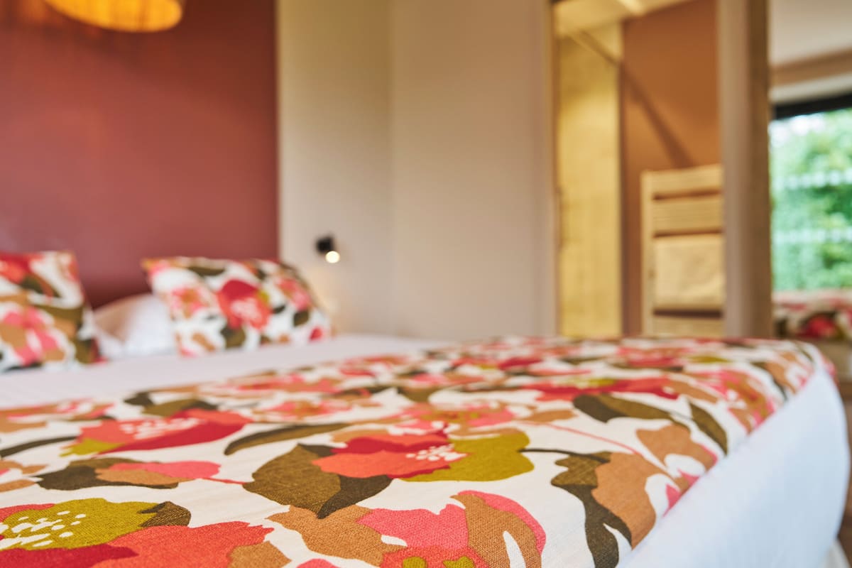 Uma cama com colcha estampada em flor, cabeceira de madeira e vista para um jardim através de uma porta aberta.