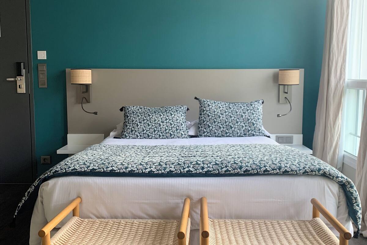 Quarto com parede verde-água, cama com colcha estampada, duas luminárias de cabeceira e um banquinho ao pé da cama.