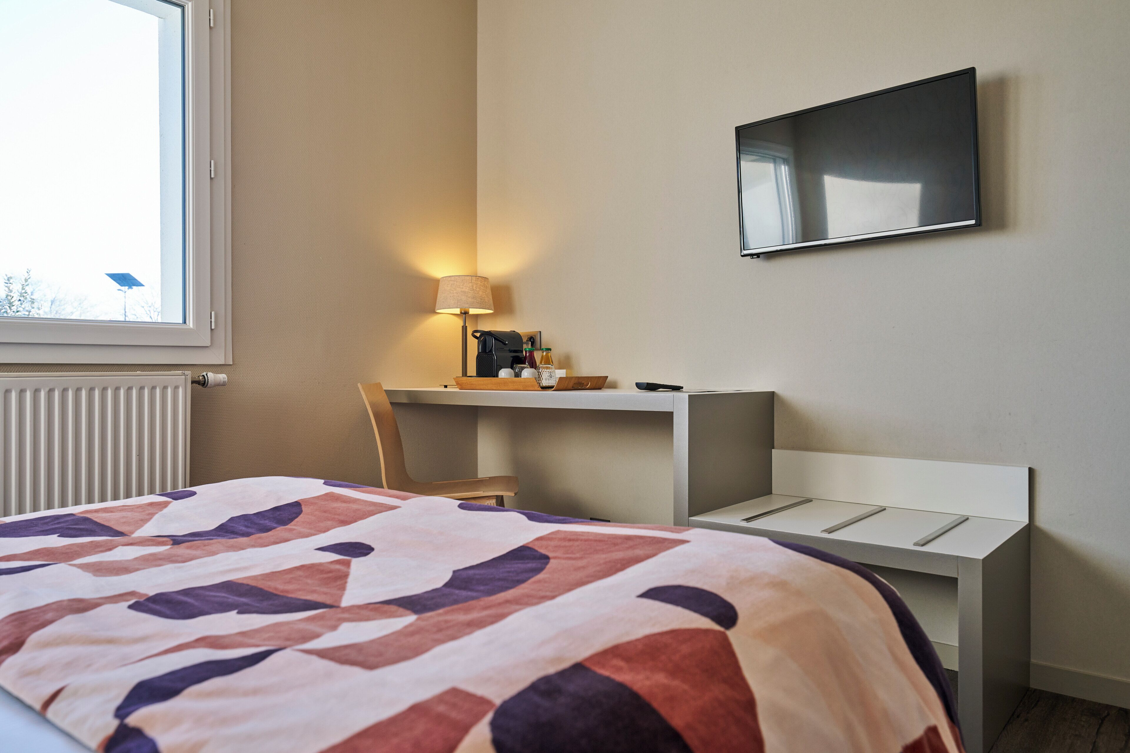 Quarto de hotel com cama, escrivaninha, cadeira, luminária e televisão na parede.