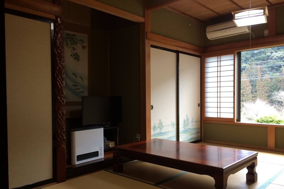 Um quarto japonês tradicional com tatames, uma mesa baixa, uma televisão, portas deslizantes e vista para uma floresta pela janela.