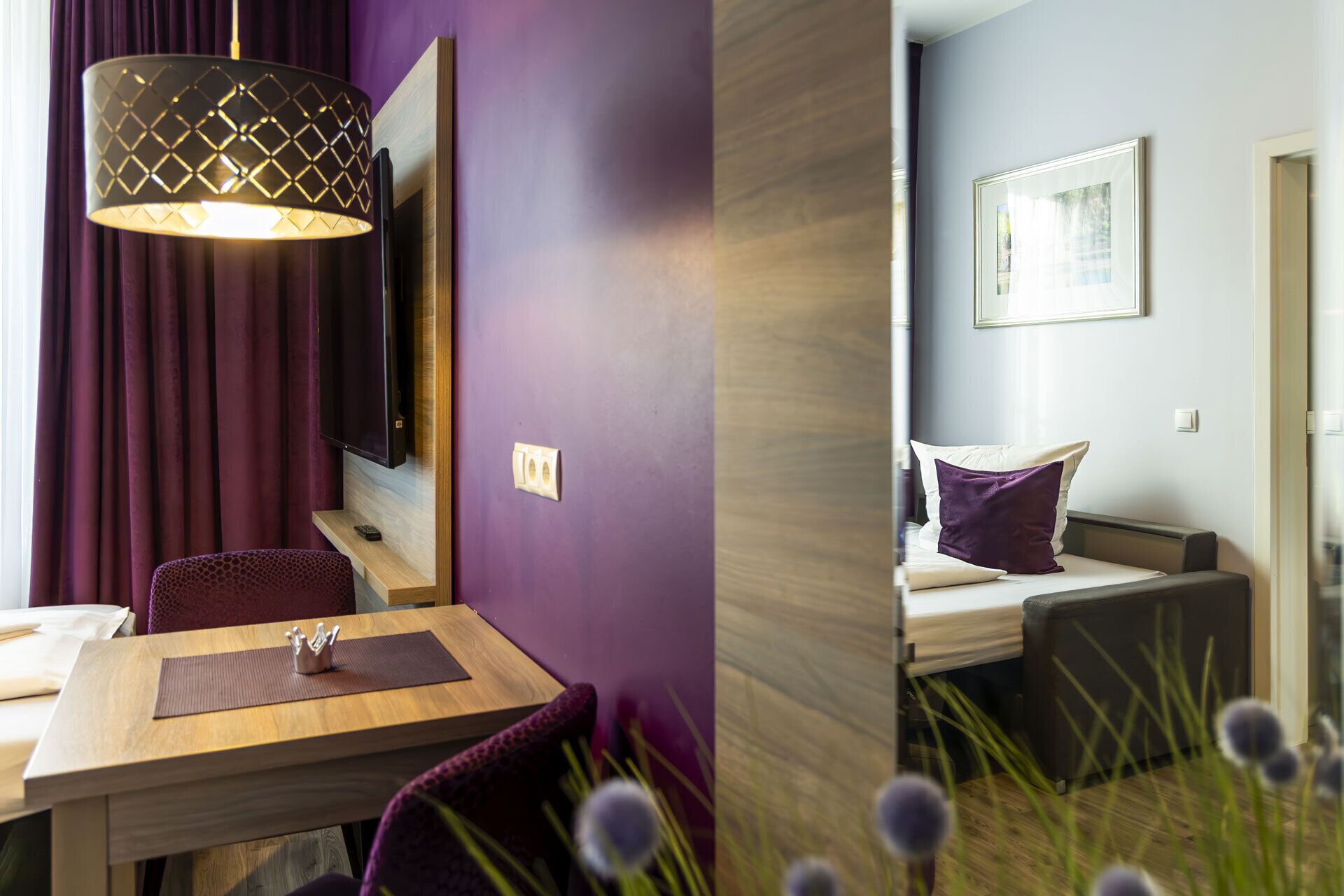 Quarto moderno com mesa de jantar, cortinas roxas, televisão na parede e cama com travesseiro roxo.