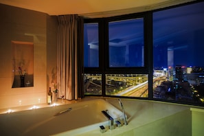 Um banheiro moderno com uma banheira grande, uma janela com vista para a cidade e um lavatório com um vaso e garrafas.
