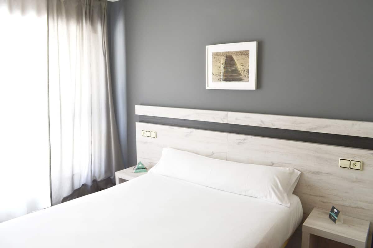 Quarto de hotel com cama, mesa de cabeceira, tomada e um quadro na parede.