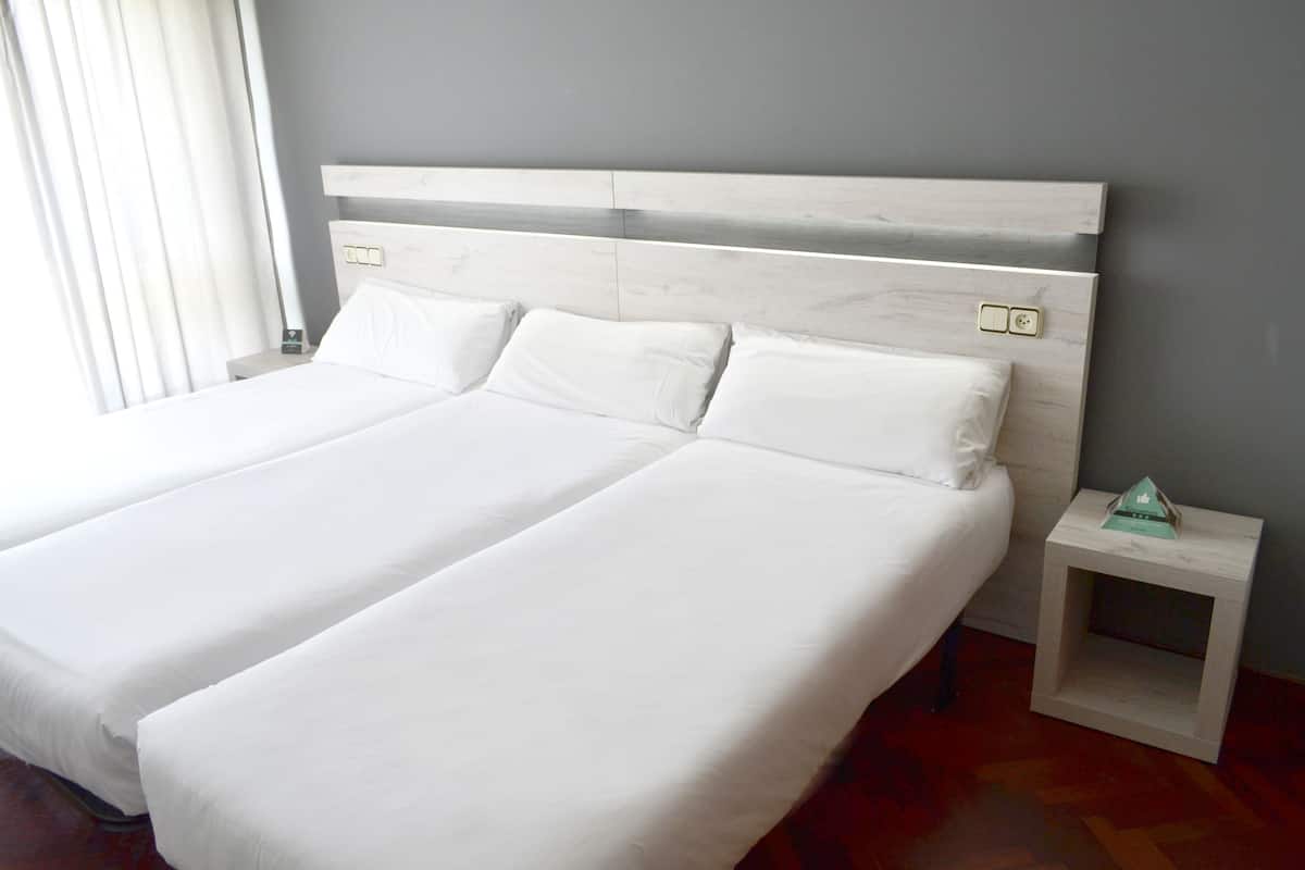 Uma cama de casal com cabeceira de madeira, uma mesa de cabeceira com abajur e vista para uma janela com cortinas.