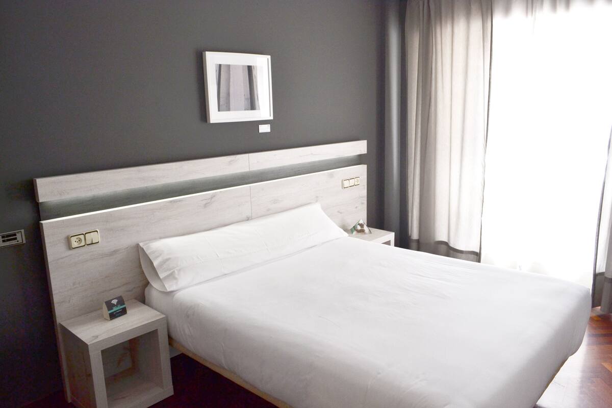 Cama bem arrumada em quarto de hotel com parede cinza, mesa de cabeceira pequena e janela com cortinas.