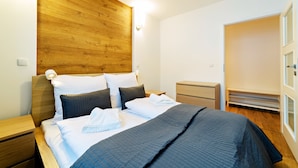 Quarto moderno com cabeceira de madeira, cama bem arrumada com roupa de cama branca e escura, mesa de cabeceira com abajur e closet espaçoso com porta espelhada.