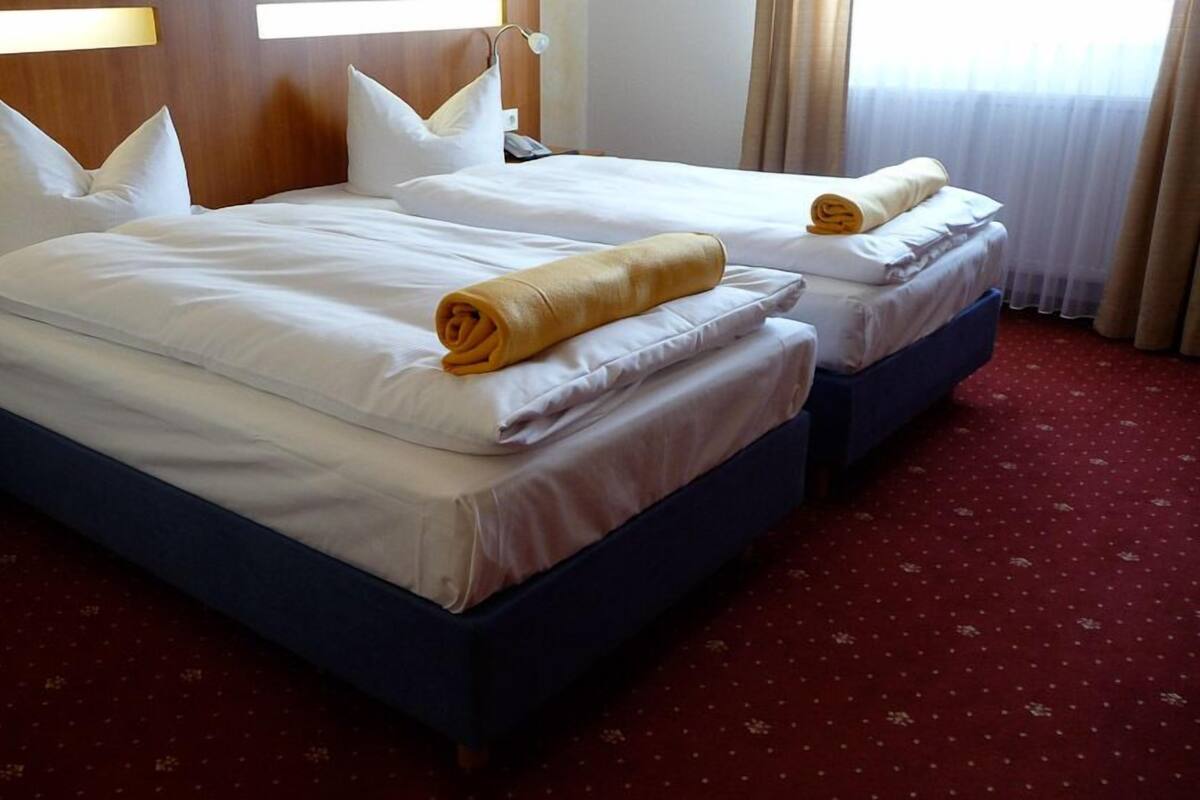 Quarto de hotel com duas camas, cada uma com lençóis e almofadas brancas, uma cabeceira de madeira, uma pequena mesa de cabeceira de madeira e um chão coberto por carpete vermelho.