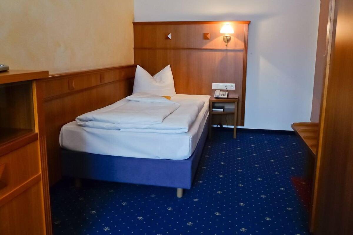 Quarto de hotel com uma cama, mesa de cabeceira com abajur e uma pequena prateleira.