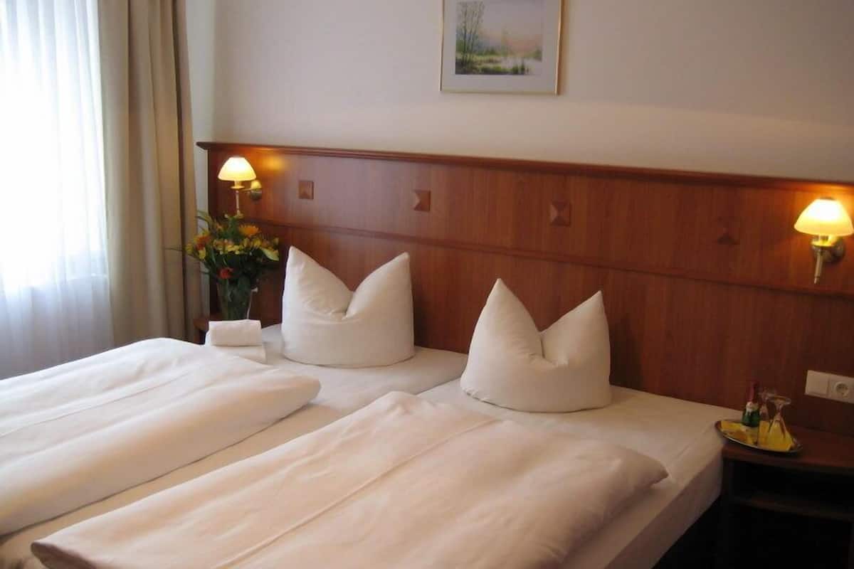 Quarto de hotel com duas camas, uma cabeceira de madeira, um quadro pendurado na parede e uma mesa de cabeceira com abajur e bandeja.