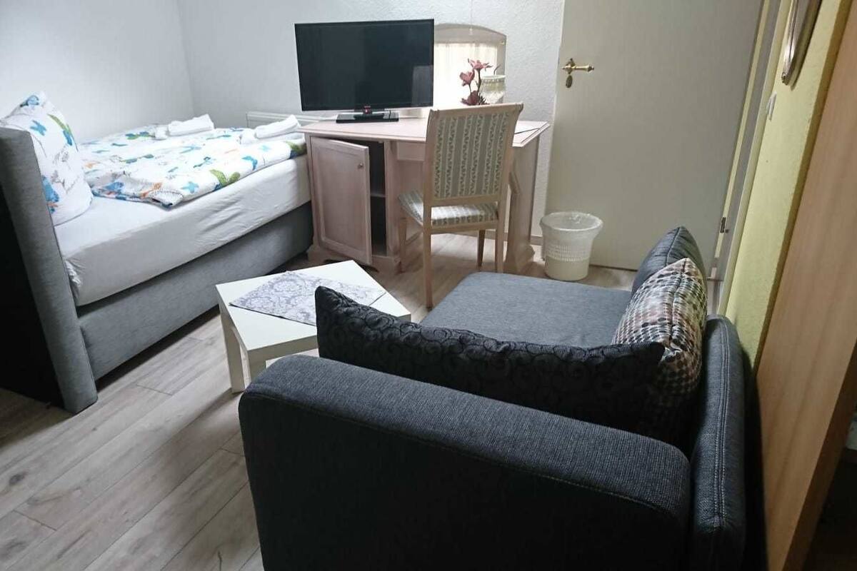 Quarto compacto com cama, escrivaninha com computador, mesa pequena e sofá.