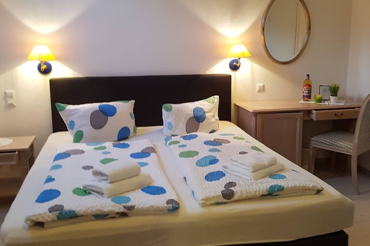 Uma cama de casal com colcha de bolinhas, duas luminárias de cabeceira, uma escrivaninha, uma cadeira e um espelho na parede.