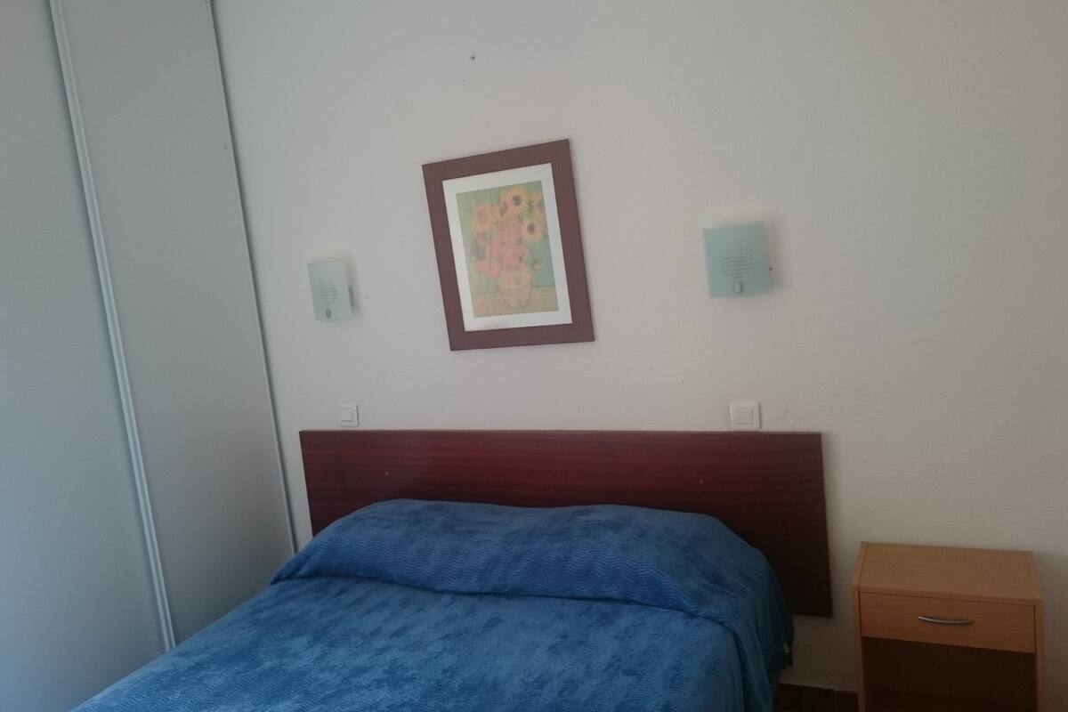 Quarto com cama, mesa de cabeceira, luminária na parede e quadro emoldurado.