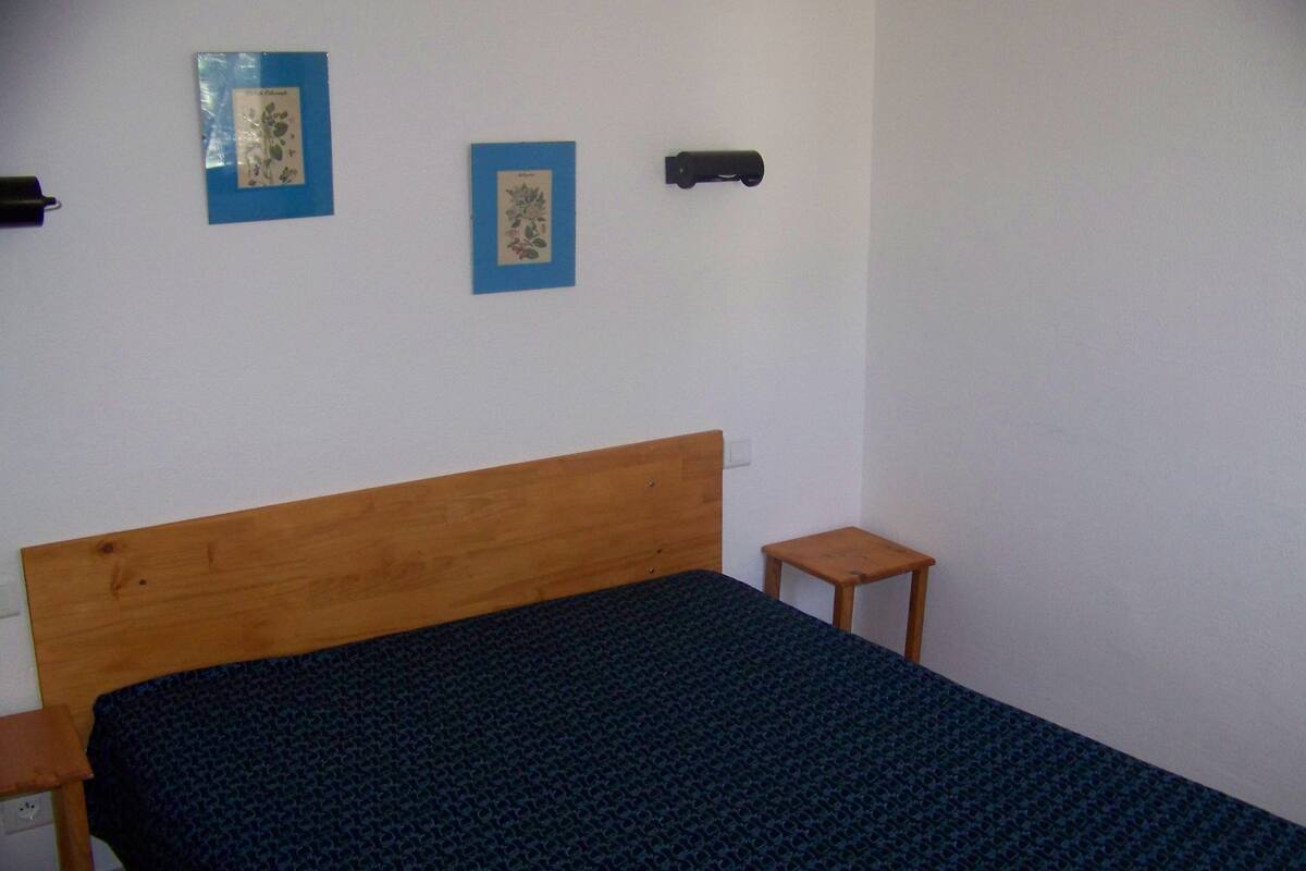 Quarto com cabeceira de madeira, uma cama de solteiro, uma mesa de cabeceira pequena de madeira e duas imagens emolduradas na parede.
