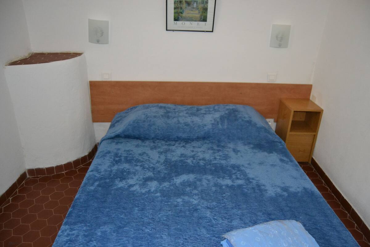 Quarto pequeno com uma cama de solteiro, colcha azul, cabeceira de madeira, mesa de cabeceira de madeira e interruptor de luz fixado na parede.