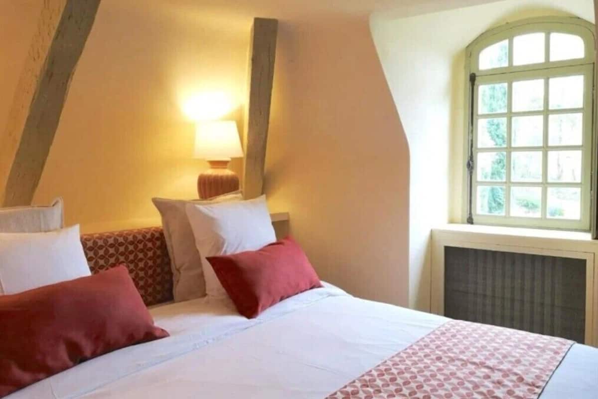 Quarto com cama, almofadas, luminária de cabeceira, janela e radiador.