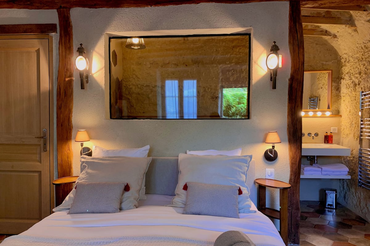 Quarto com cama grande, duas luminárias de cabeceira, espelho sobre a cama, porta de madeira e banheiro com pia e espelho.
