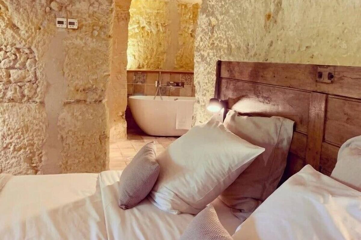 Quarto com paredes de pedra, uma cabeceira de madeira, roupa de cama branca e uma banheira ao fundo.