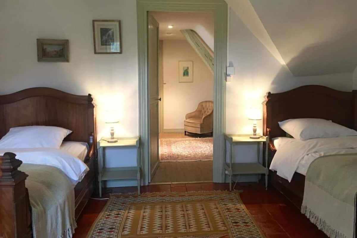 Quarto com duas camas, cabeceira de madeira, mesa de cabeceira com abajur, uma cadeira ao fundo e porta que dá para outro cômodo.