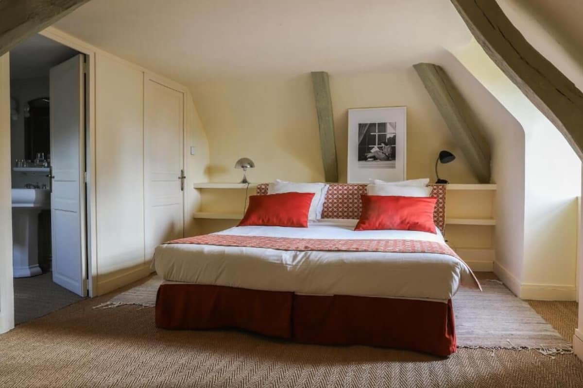 Quarto com cama grande, teto de madeira e pequena mesa de cabeceira com luminária.