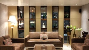 Uma sala de estar moderna com um sofá de couro marrom, duas poltronas, uma mesa de centro e um abajur. Há várias prateleiras com objetos decorativos e plantas.