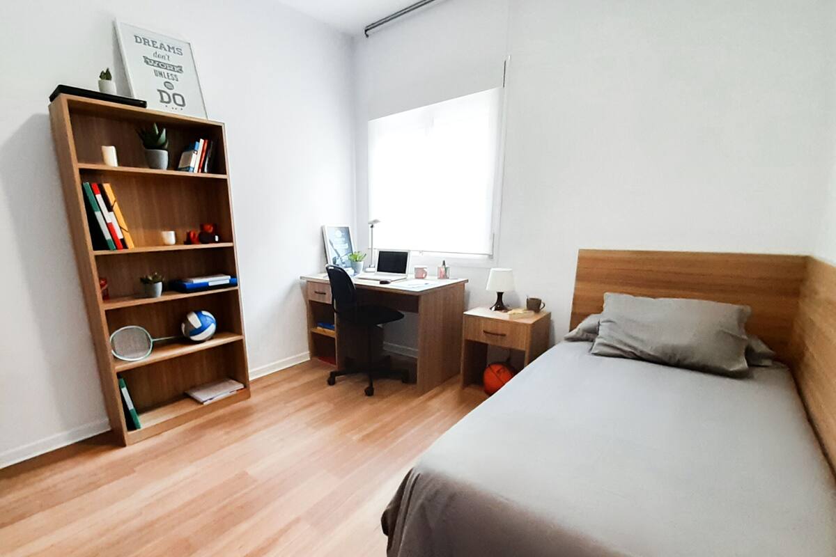 Quarto com cama, escrivaninha, computador, estante e janela.