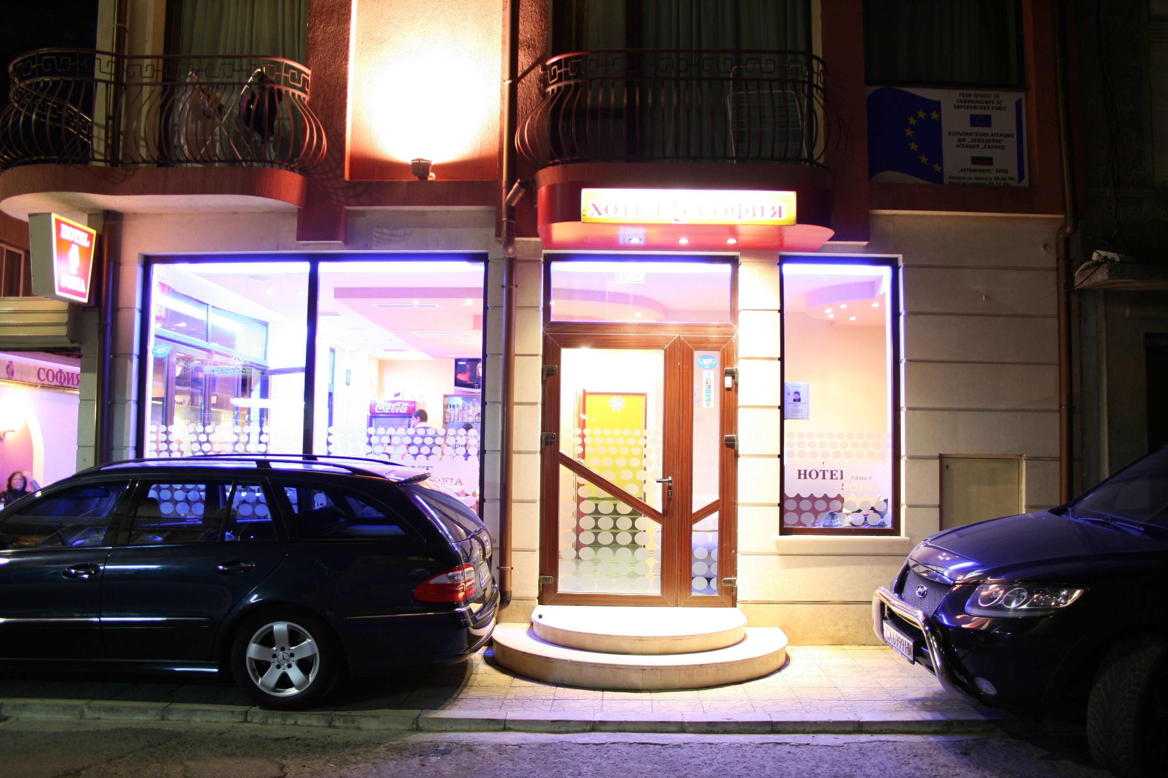 Entrada de hotel com uma placa bem iluminada, escada que leva à entrada e um carro estacionado na frente.