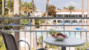 Varanda com mesa, exibindo uma tigela de frutas, com vista para área da piscina, espreguiçadeiras e palmeiras.