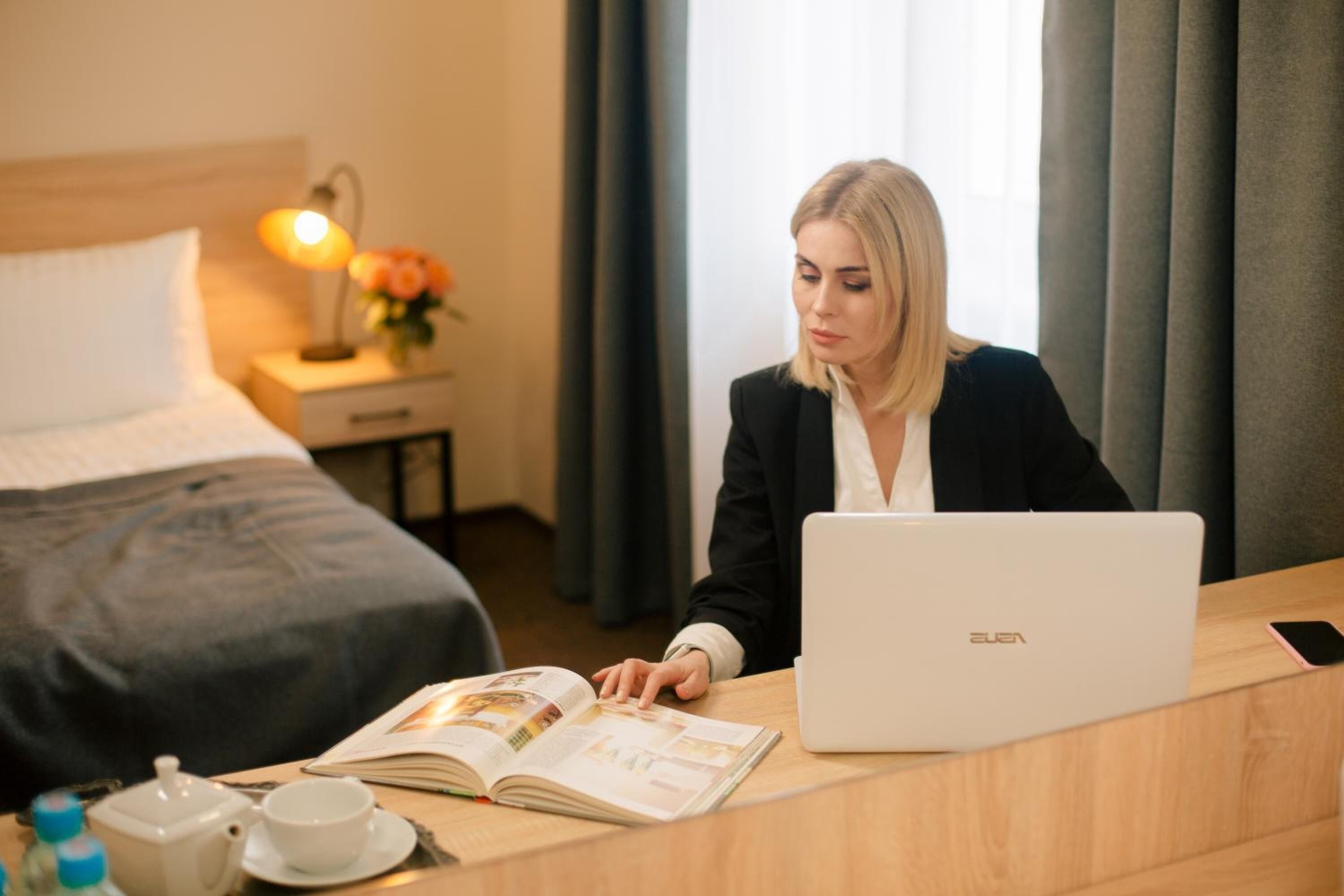Quarto de hotel com cama, mesa de cabeceira com abajur, um laptop, um livro, uma chávena e um bule de chá.