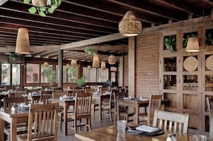 Restaurante com mesas e cadeiras de madeira, luminárias de tecido penduradas e parede de madeira com prateleiras.