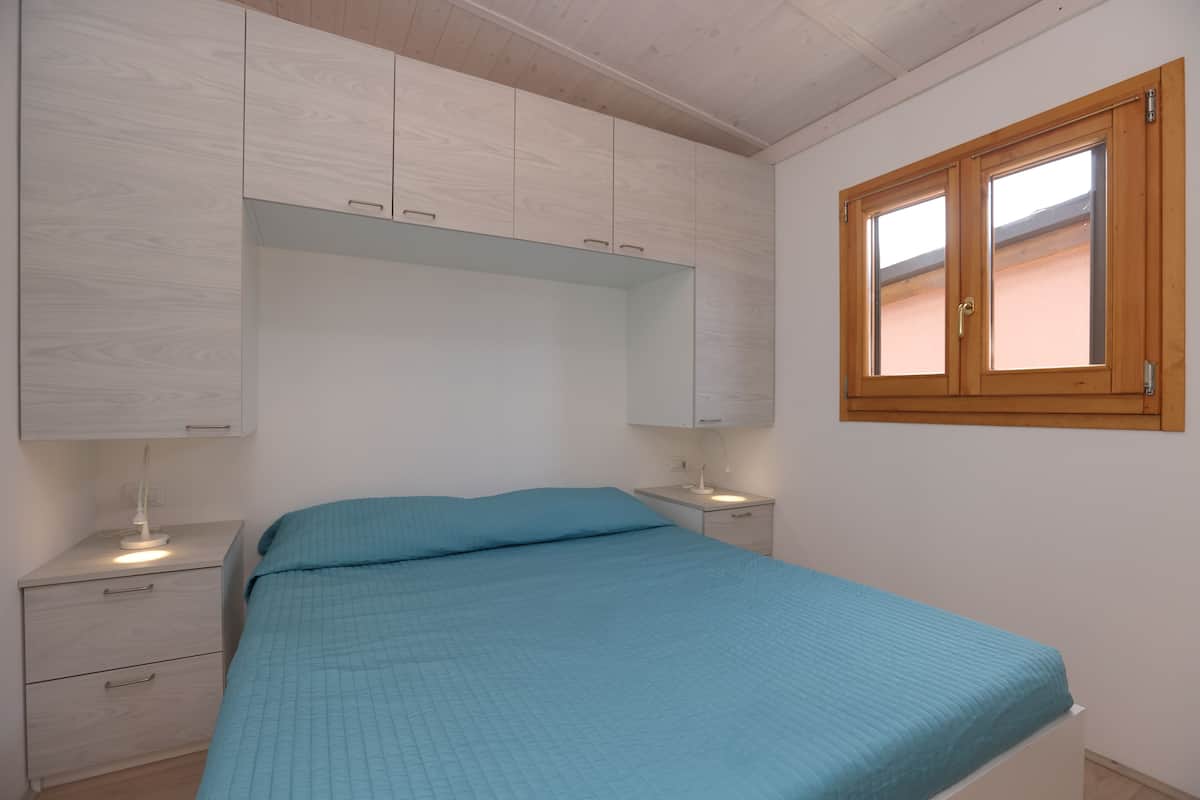 Quarto com cama, mesa de cabeceira, maleiro e janela com persianas de madeira.