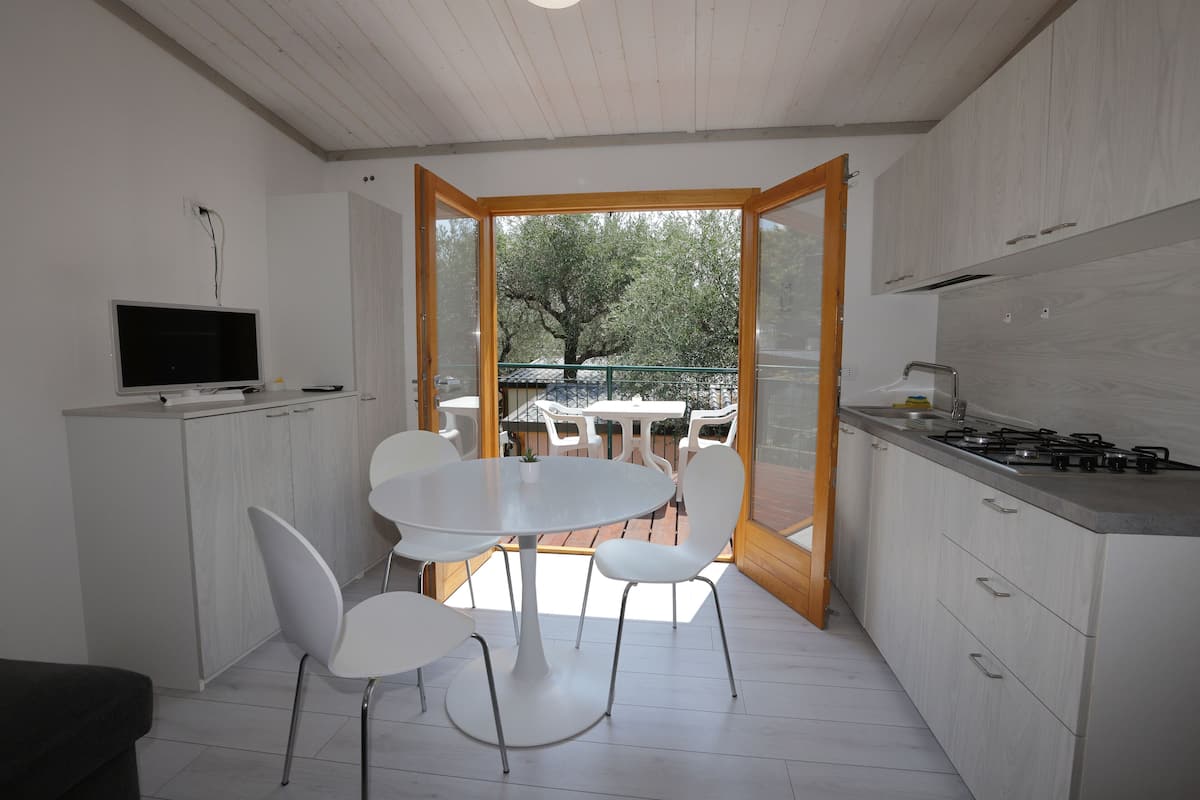Cozinha moderna com armários brancos, área de jantar com mesa e cadeiras redondas, e vista para árvores através de uma porta aberta.