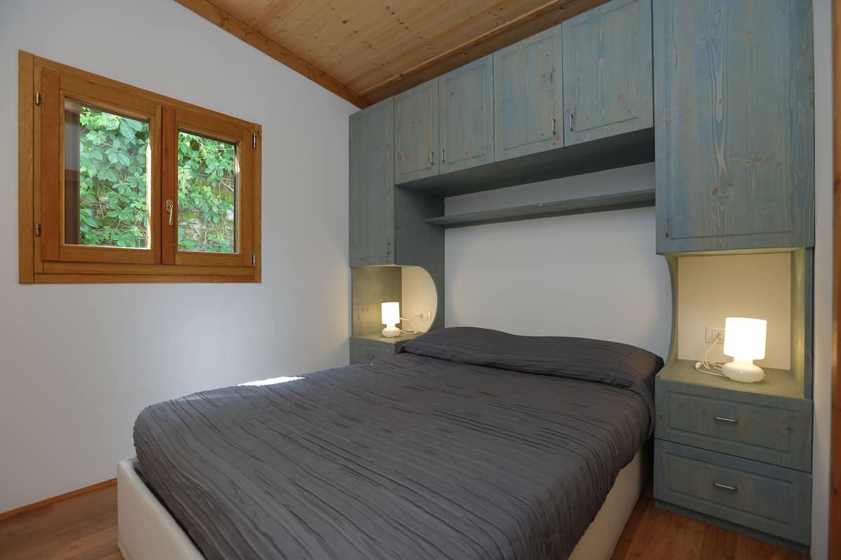 Quarto com cama, armários de madeira, mesa de cabeceira com abajur e janela com vegetação lá fora.